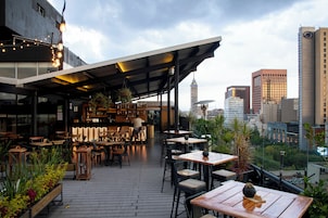 Uma área de estar ao ar livre com mesas e cadeiras, um bar com prateleiras cheias de garrafas e um horizonte urbano ao fundo.