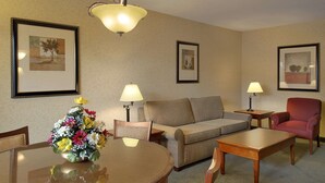 Quarto de hotel com mesa de jantar redonda, um sofá, uma poltrona vermelha e duas luminárias. Quadros emoldurados decoram as paredes.