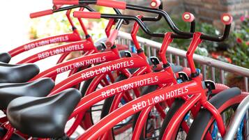 Uma fileira de bicicletas vermelhas com a marcação 'RED LION HOTELS', estacionadas em área externa com grades de metal e vegetação ao fundo.