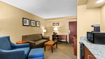 Quarto de hotel com sofá, poltrona, mesa de centro, escrivaninha, cadeira, luminária, micro-ondas e uma pequena cozinha compacta.