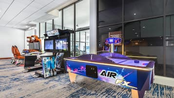 Uma mesa de bilhar com a marca 'AIR FX', uma mesa de bilhar e uma mesa de pingue-pongue em um espaço com grandes janelas e piso coberto de carpete.