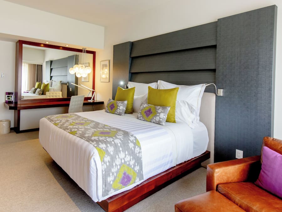 Quarto de hotel moderno com uma cama grande, cabeceira de madeira, um espelho, uma escrivaninha e uma poltrona de couro.