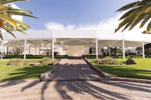 Uma galeria coberta conduz a um edifício com design arquitetônico moderno, cercado por jardins bem cuidados e palmeiras.