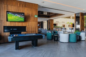 Espaço lounge moderno com mesa de bilhar, assentos confortáveis, televisão exibindo um evento esportivo e uma saída identificada.