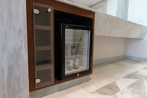 Minibar com porta de vidro, contendo um refrigerador de bebidas, uma garrafa de Corona Extra e uma lata de Pepsi.