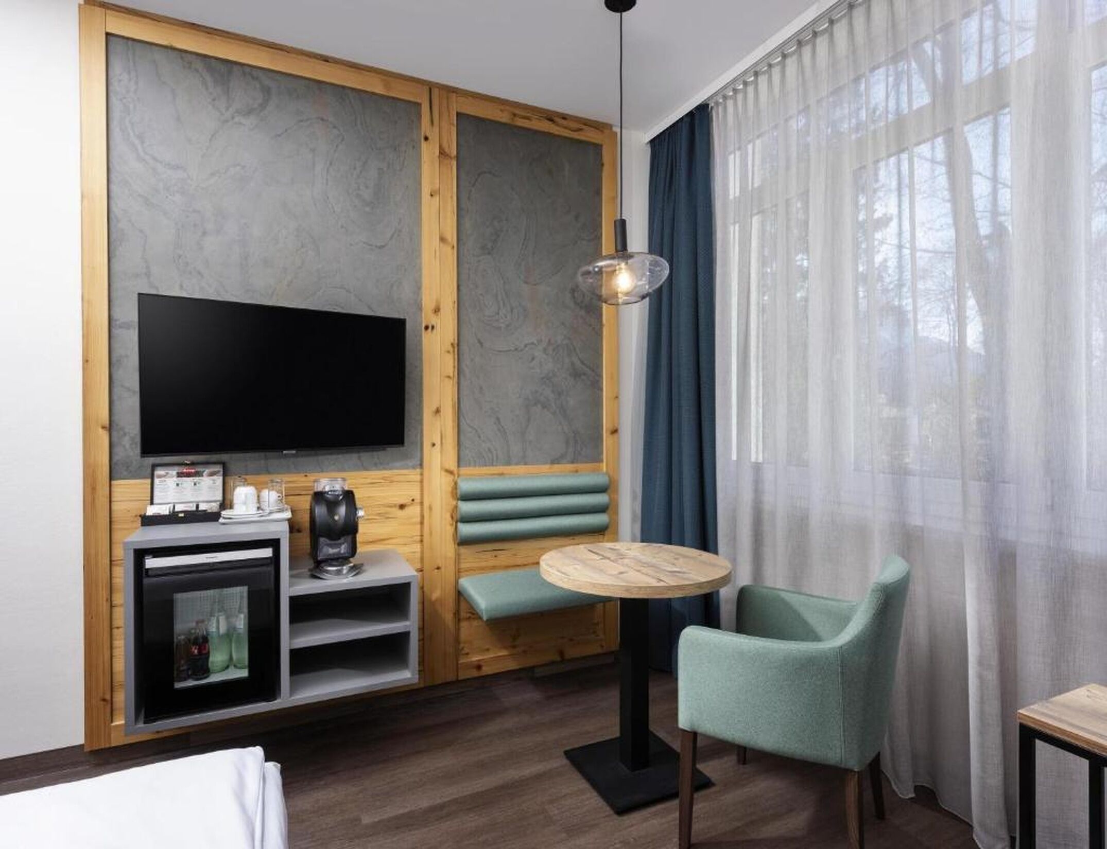 Quarto de hotel com televisão de tela plana, cafeteira, minibar, sofá com estrutura de madeira, mesa redonda e uma cadeira.