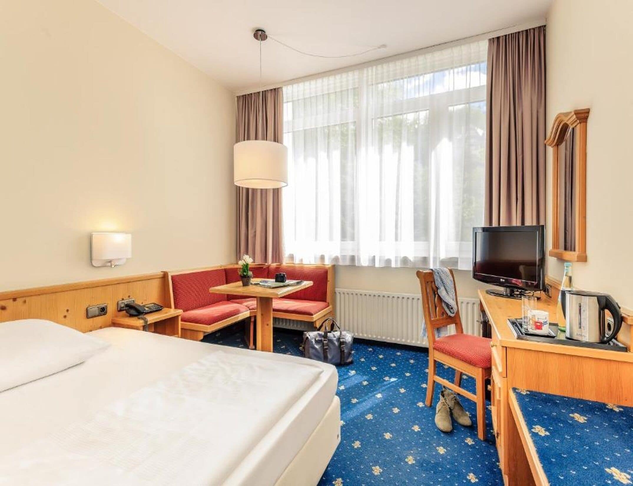 Quarto de hotel com cama, escrivaninha com computador, cadeira, mesa pequena e televisão.