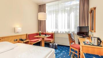 Quarto de hotel com cama, escrivaninha com computador, cadeira, mesa pequena e televisão.