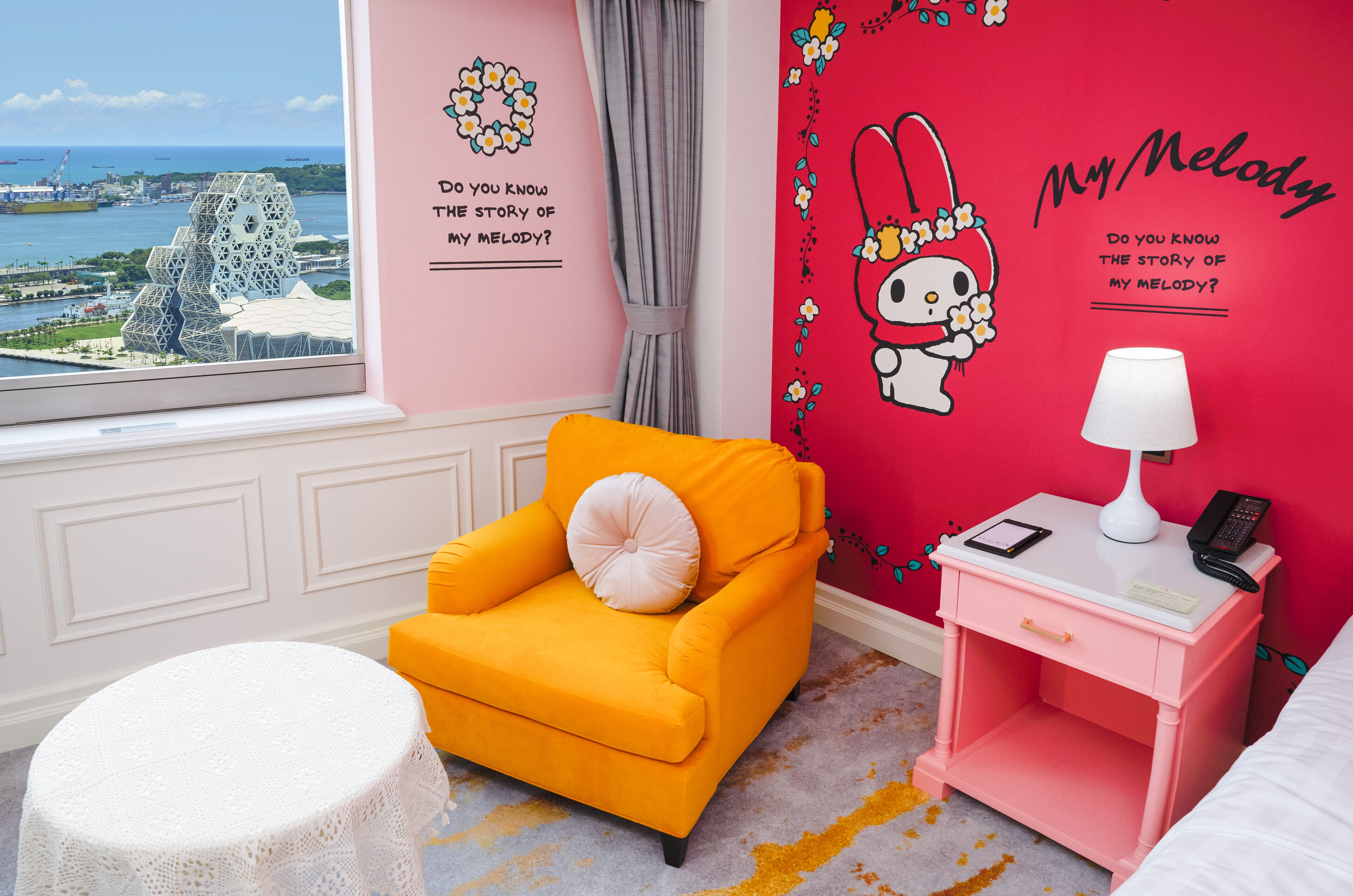 Quarto com vista para um horizonte costeiro, parede rosa com um personagem de desenho animado, poltrona amarela, mesa branca e mesa de cabeceira rosa.