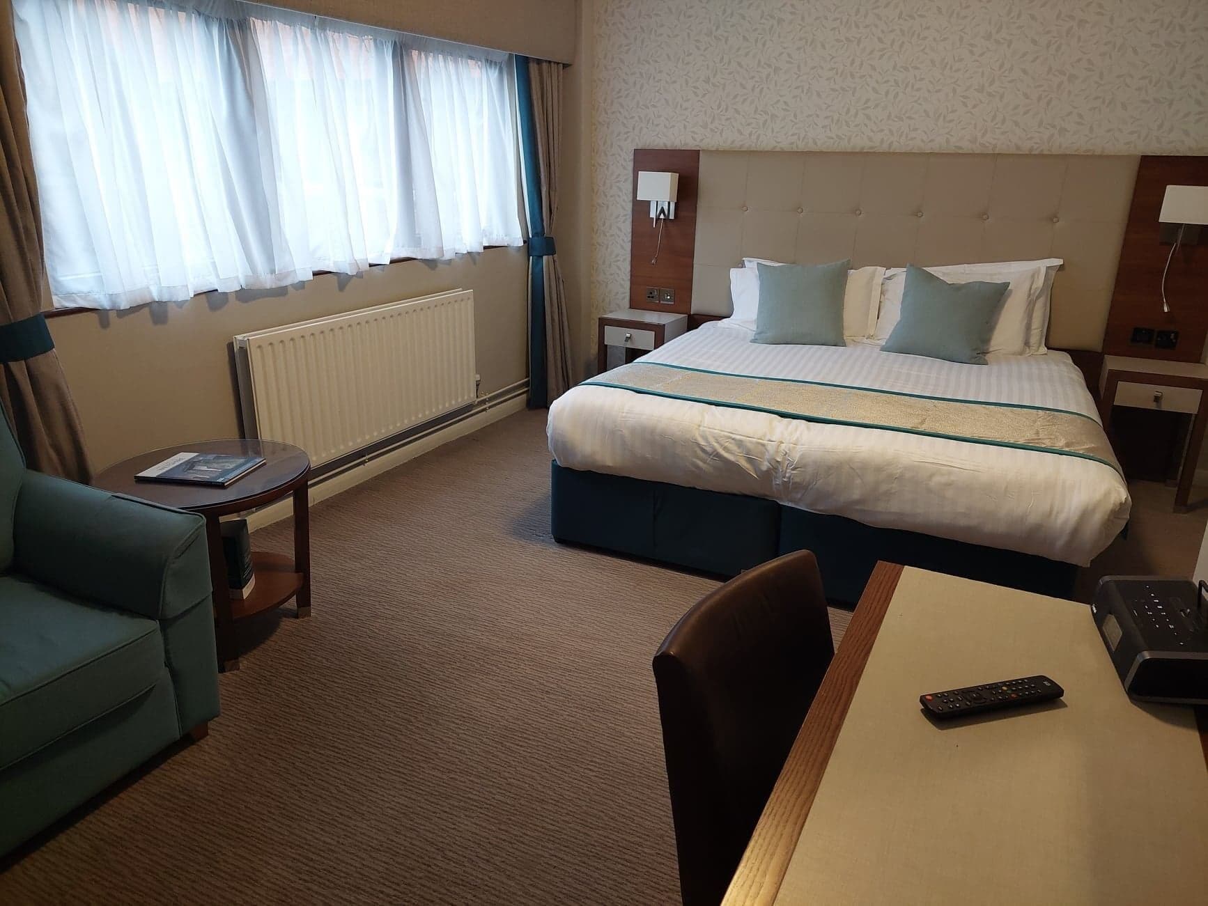 Quarto de hotel com cama, escrivaninha, cadeira, mesa, luminária, mesa de cabeceira e janela com cortinas.