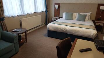 Quarto de hotel com cama, escrivaninha, cadeira, mesa, luminária, mesa de cabeceira e janela com cortinas.