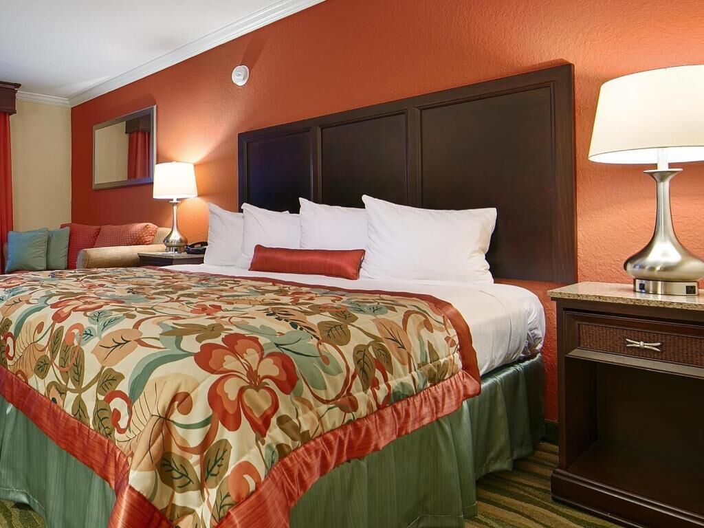 Quarto de hotel com uma cama grande, mesa de cabeceira, luminária, cômoda e espelho na parede.