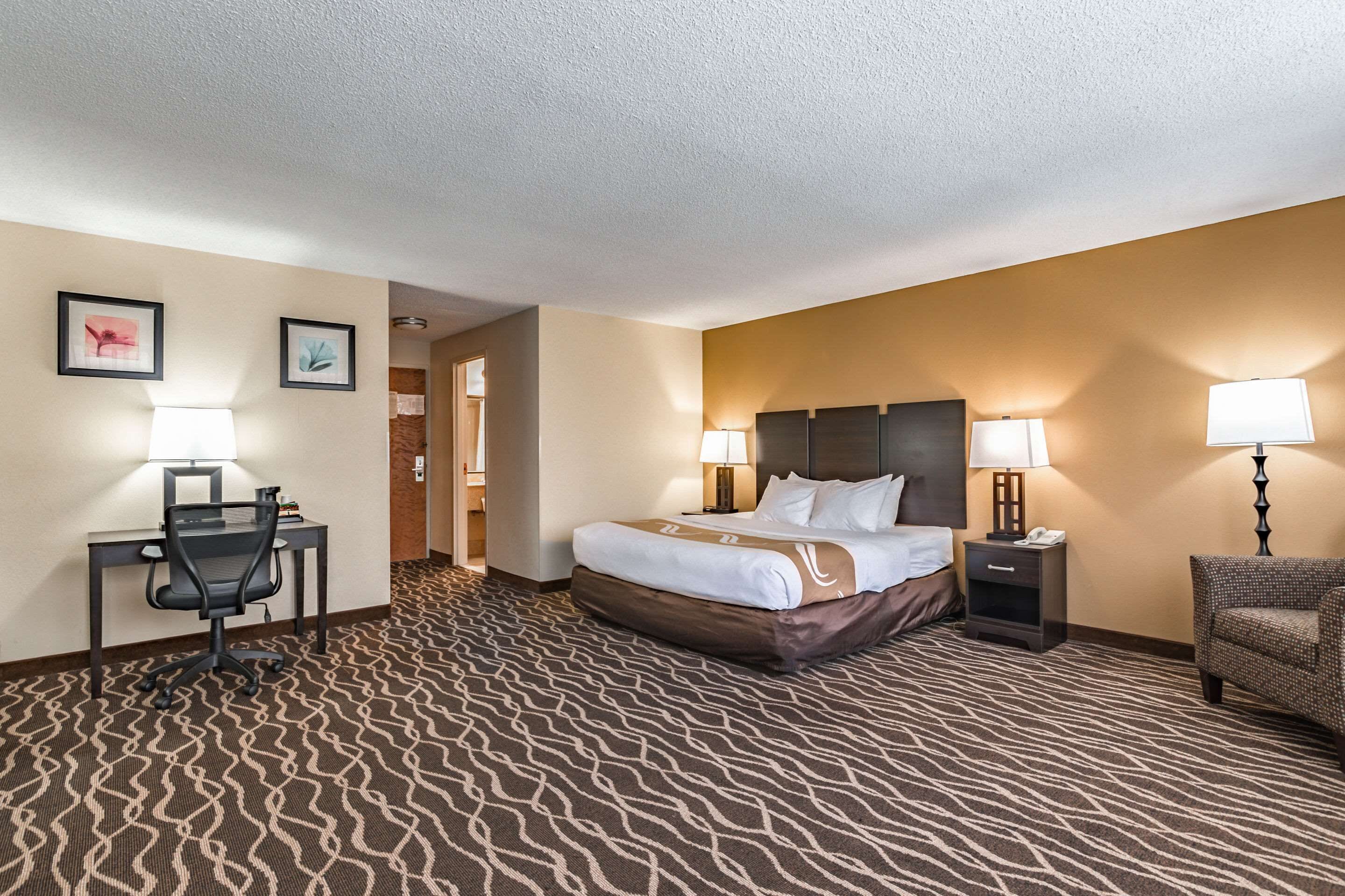 Quarto de hotel com cama, escrivaninha, cadeira, duas luminárias de cabeceira e um tapete estampado.