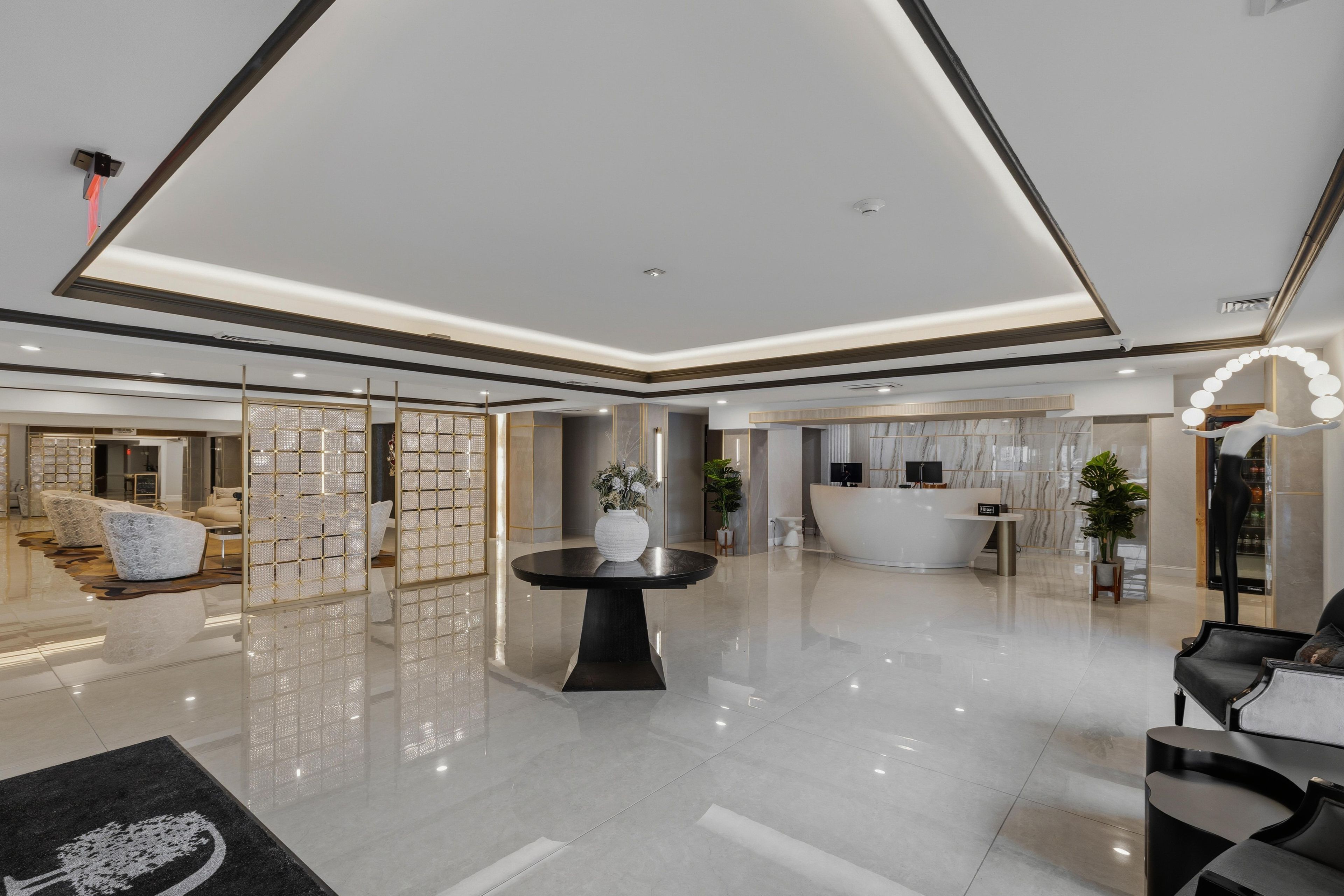 Um hall amplo com uma mesa redonda central, balcão de recepção e área de estar com sofás.