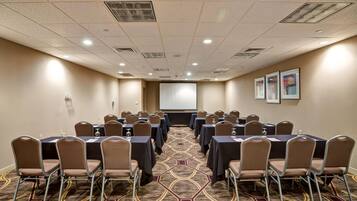 Sala de conferências com mesas, cadeiras, tela de projeção e piso coberto de carpete.