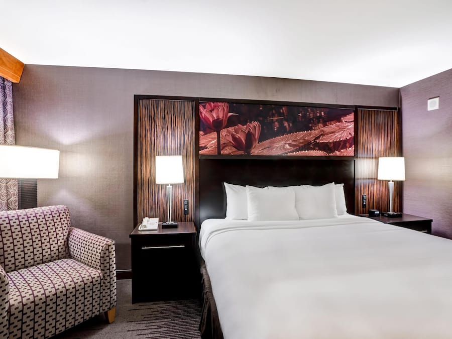 Quarto de hotel com uma cama grande, duas mesas de cabeceira, uma cadeira e uma televisão fixada na parede.