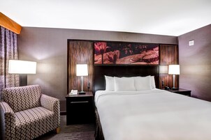 Quarto de hotel com uma cama grande, duas mesas de cabeceira, uma cadeira e uma televisão fixada na parede.