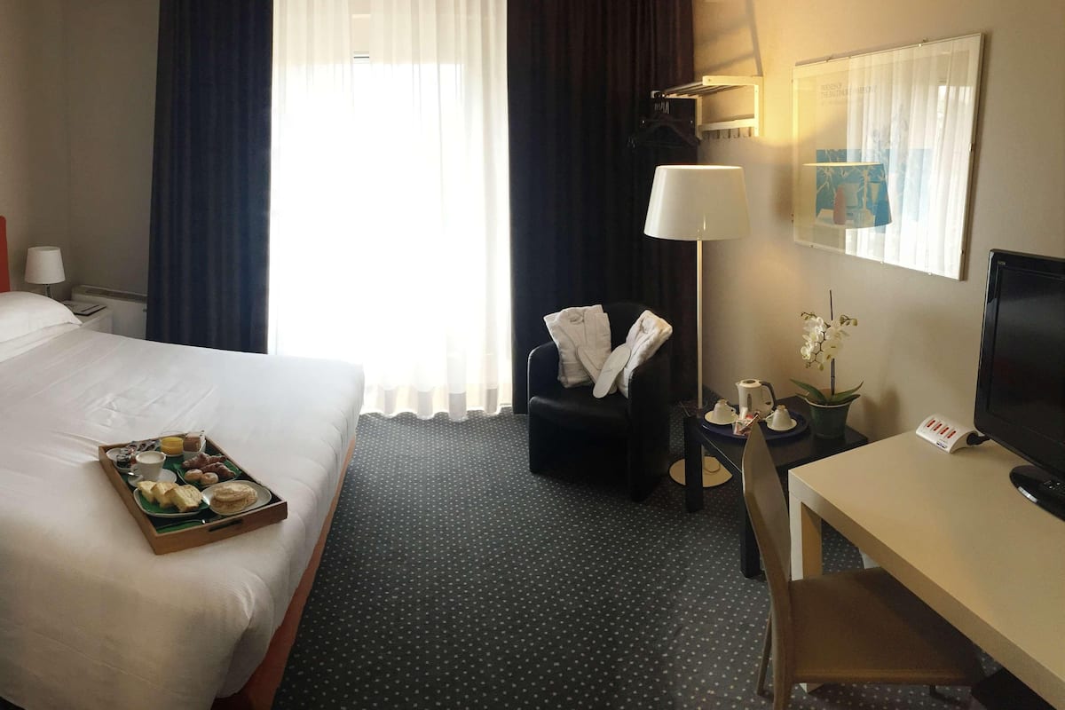Quarto de hotel com cama, escrivaninha, cadeira, bandeja com itens de café da manhã e janela com cortinas.