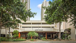Entrada de hotel com área externa coberta, palmeiras e vista para edifícios altos ao fundo.