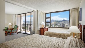 Quarto de hotel com duas camas, varanda, uma cadeira e vista para a cidade e montanhas.
