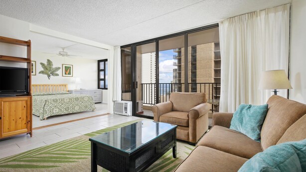 Uma sala de estar com sofá, mesa de centro, televisão, cama, varanda com vista para edifícios e ventilador de teto.