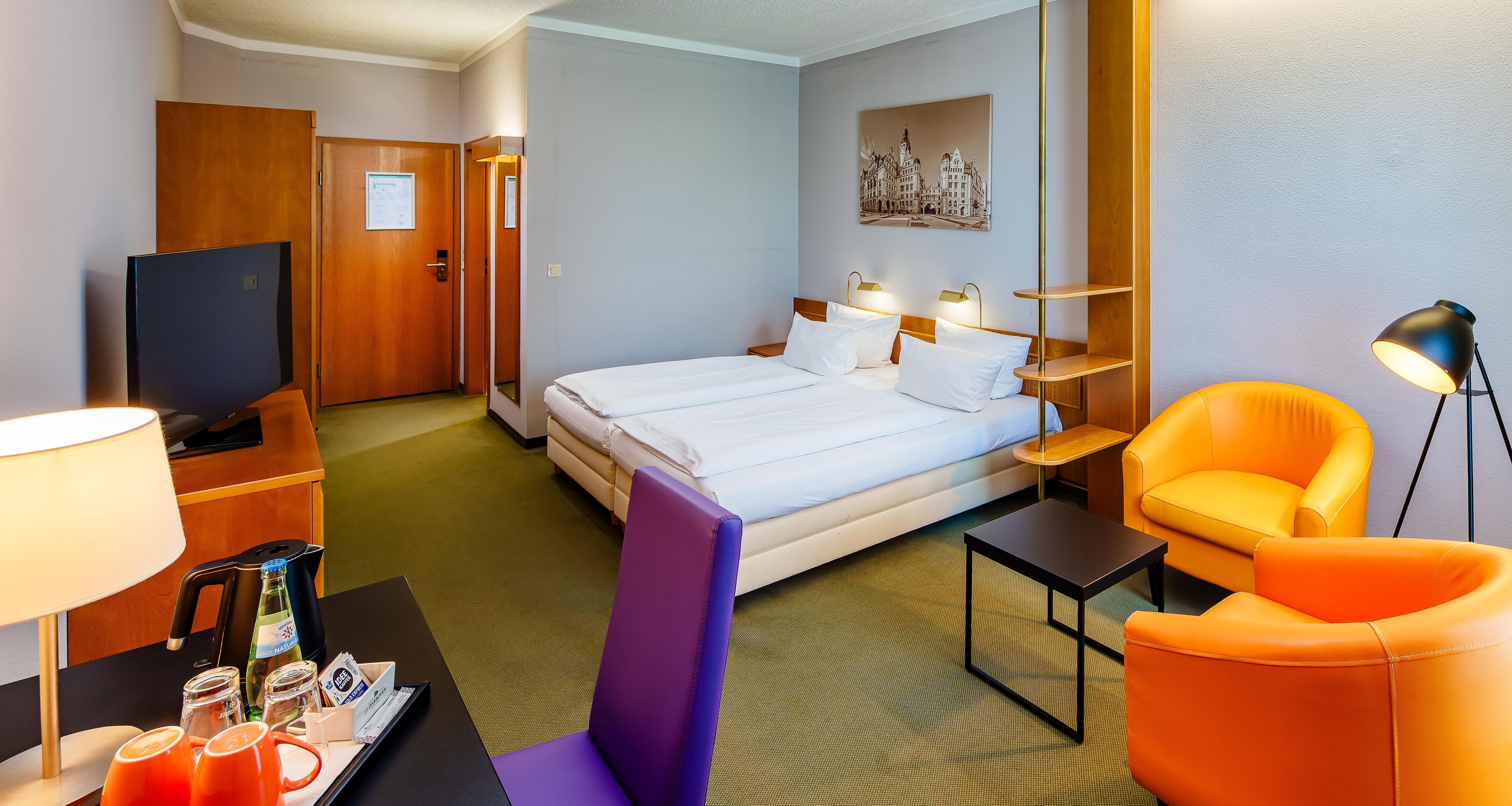 Quarto de hotel com uma cama, televisão, escrivaninha com luminária, uma cadeira e uma mesinha.