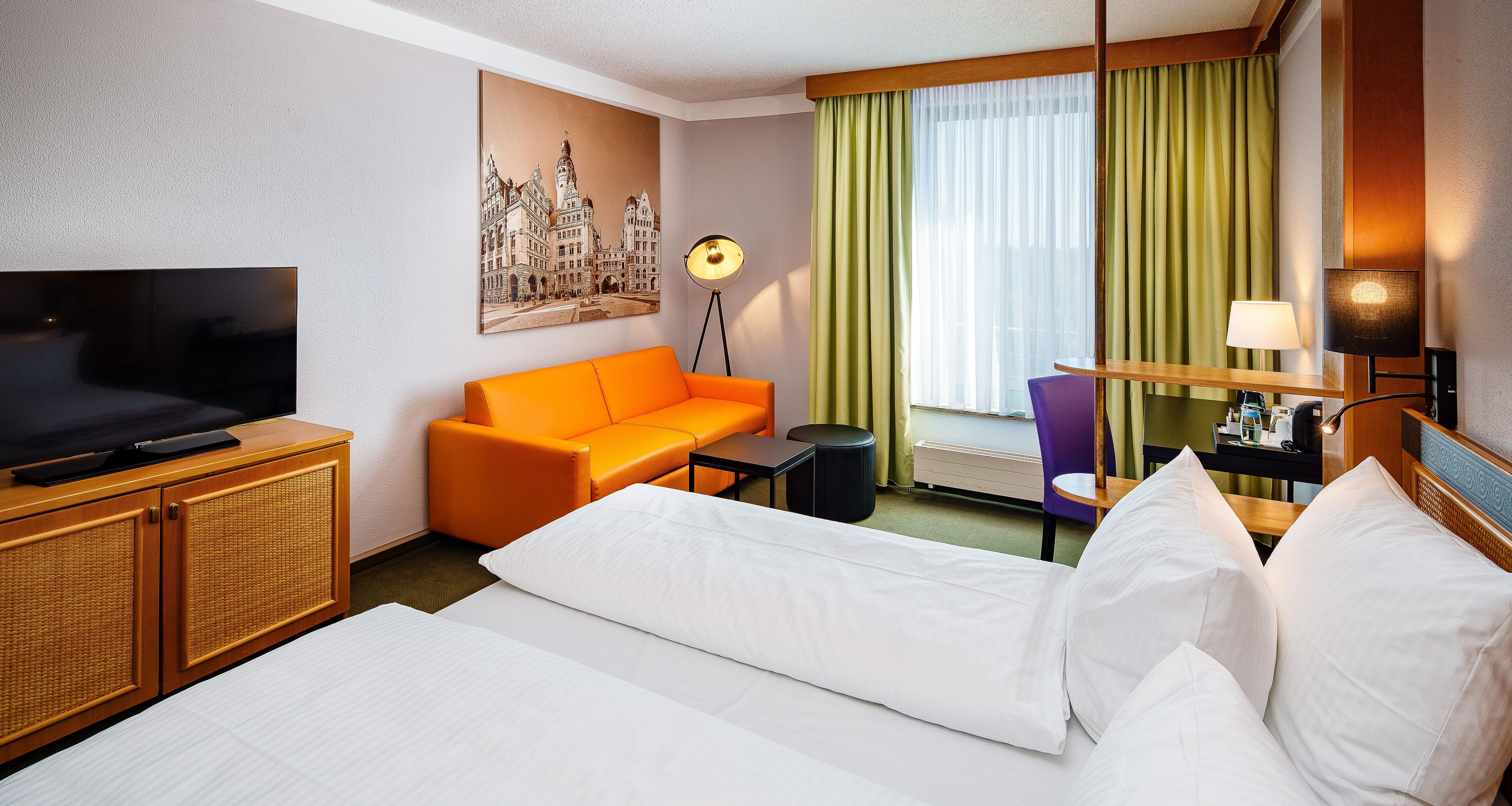 Quarto de hotel com cama, sofá, televisão, abajur e janela com cortinas.