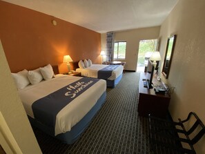 Quarto de hotel com duas camas, uma escrivaninha, uma cadeira, uma televisão e janela com vista para o verde.