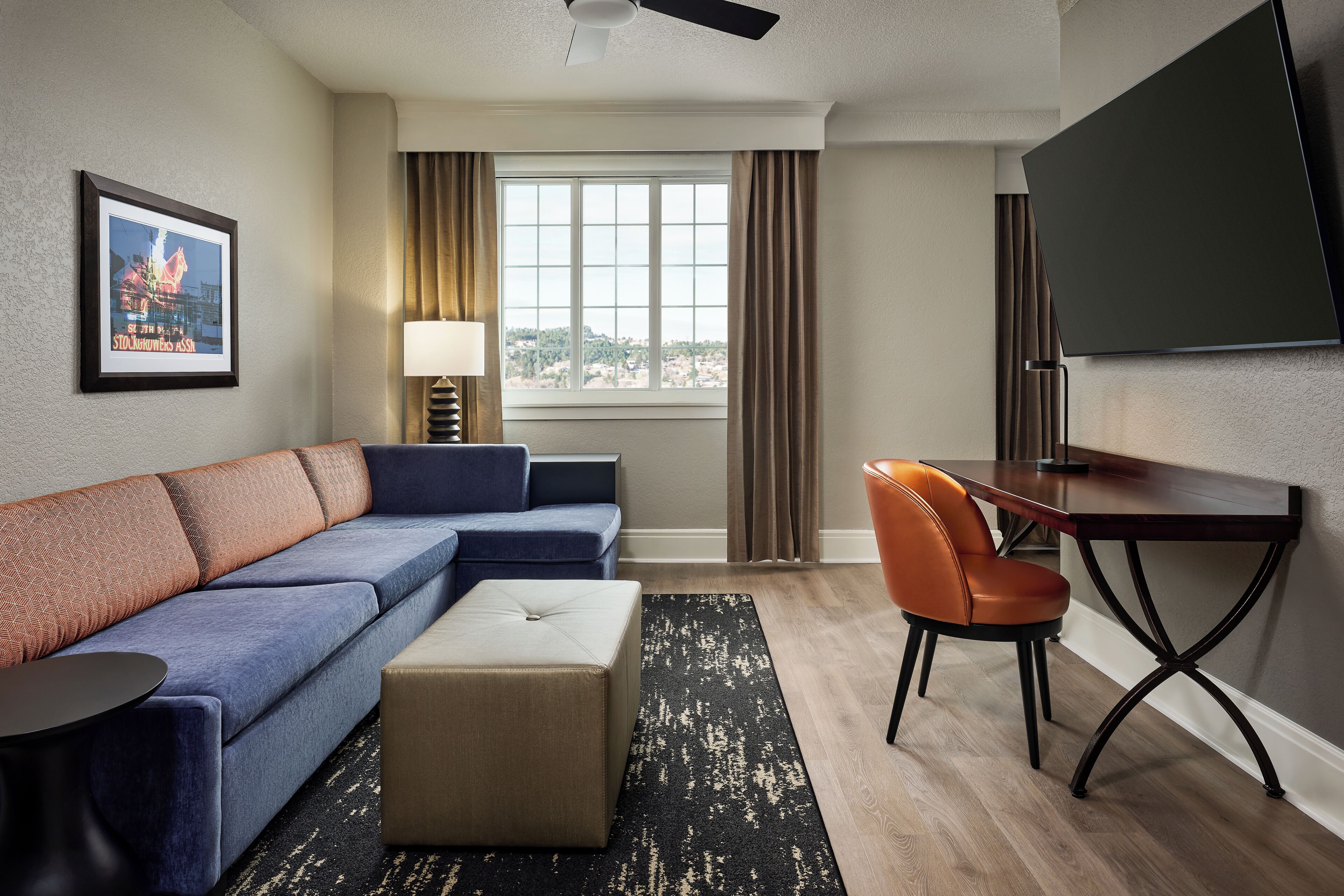 Quarto de hotel com sofá seccional, um pequeno banquinho, escrivaninha com cadeira, televisão de tela plana, janela com cortinas e luminária.
