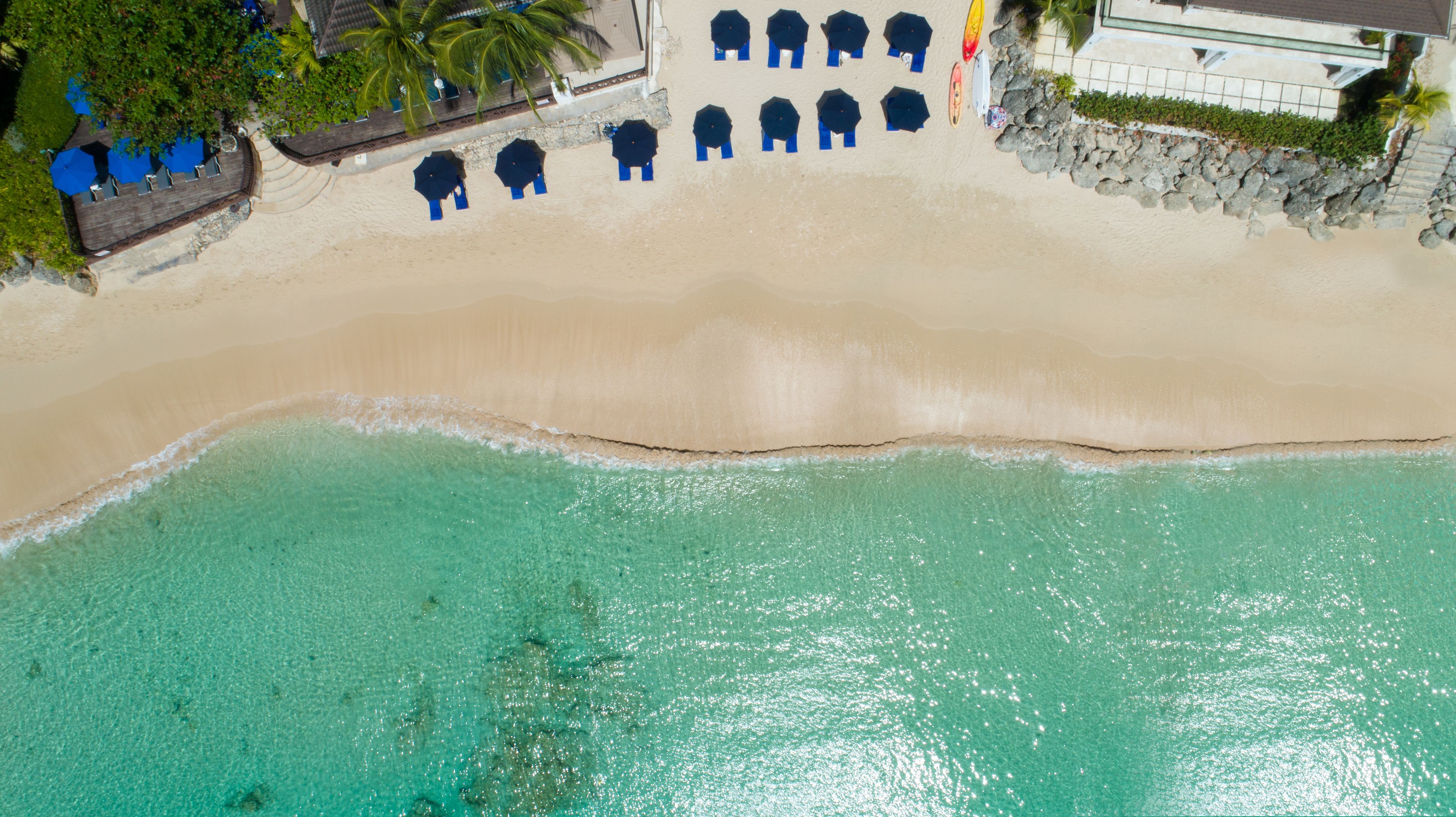 Vista aérea de uma praia com águas turquesa cristalinas, areal arenoso e vários guarda-sóis dispostos na orla.