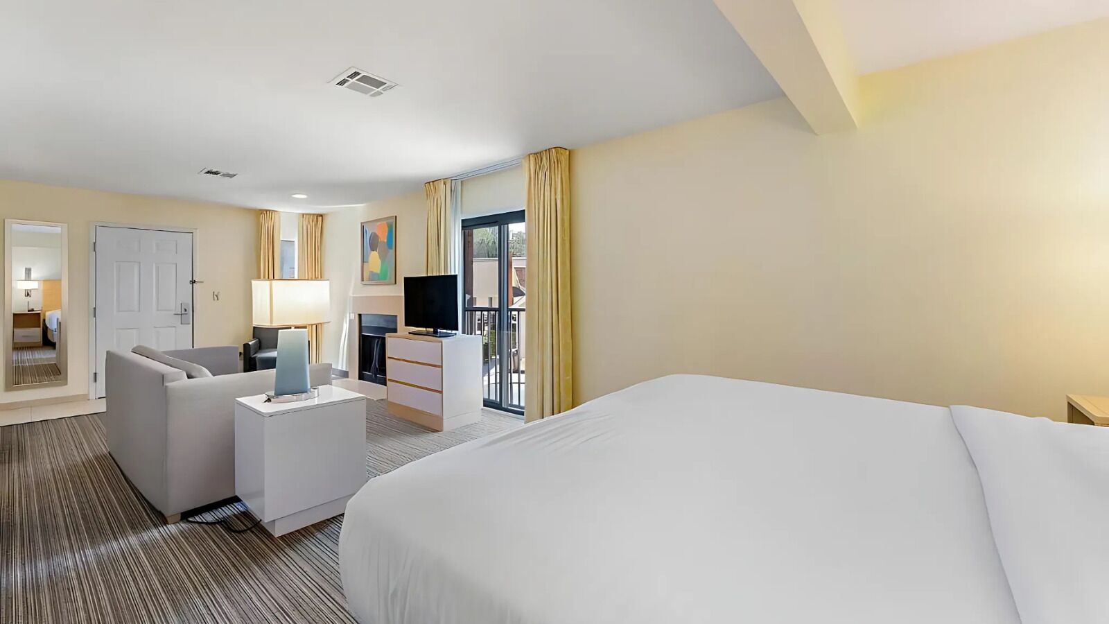 Quarto de hotel com cama, escrivaninha com cadeira, televisão e uma janela grande com cortinas.