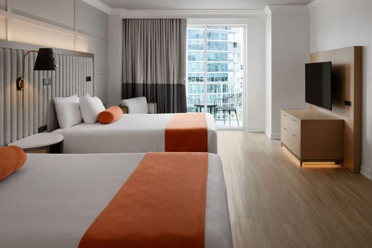 Quarto de hotel com duas camas, televisão, escrivaninha e varanda com vista para a cidade.
