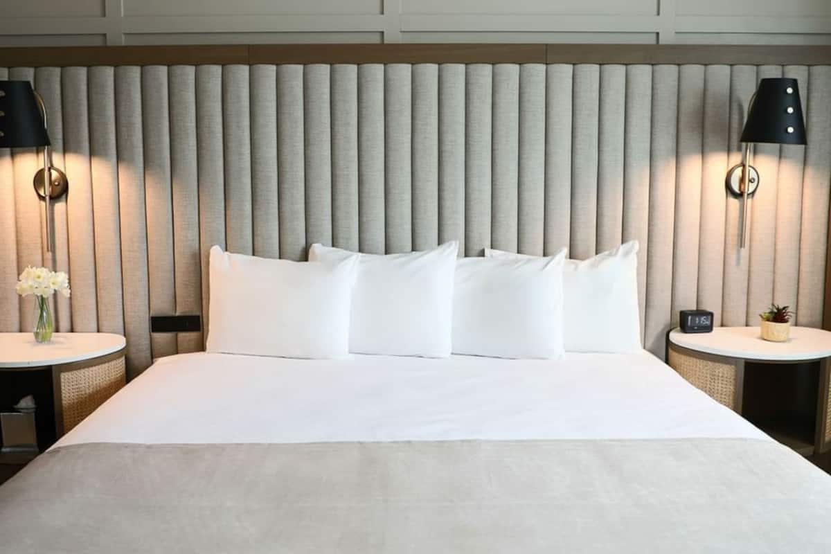 Quarto de hotel com uma cama grande, duas mesas de cabeceira, um encosto e duas luminárias fixadas na parede.