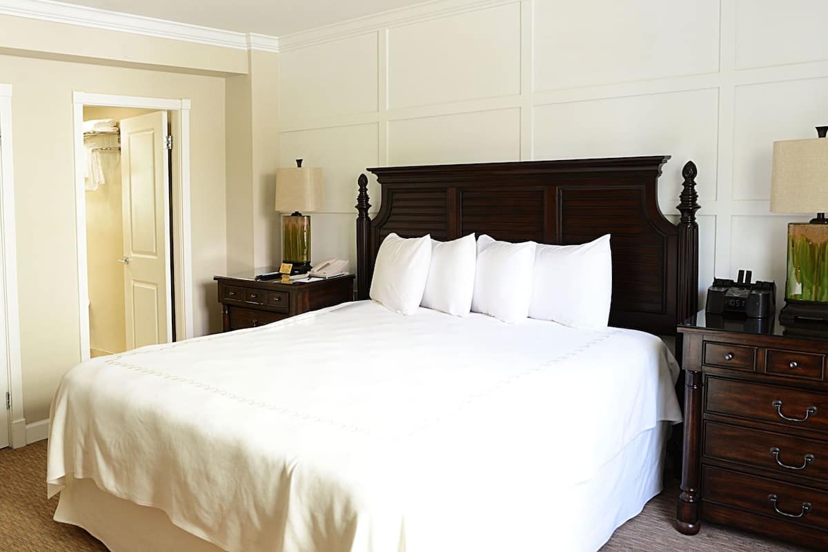 Cama bem arrumada com lençóis brancos, cabeceira de madeira, mesa de cabeceira com abajur e cômoda com gavetas.