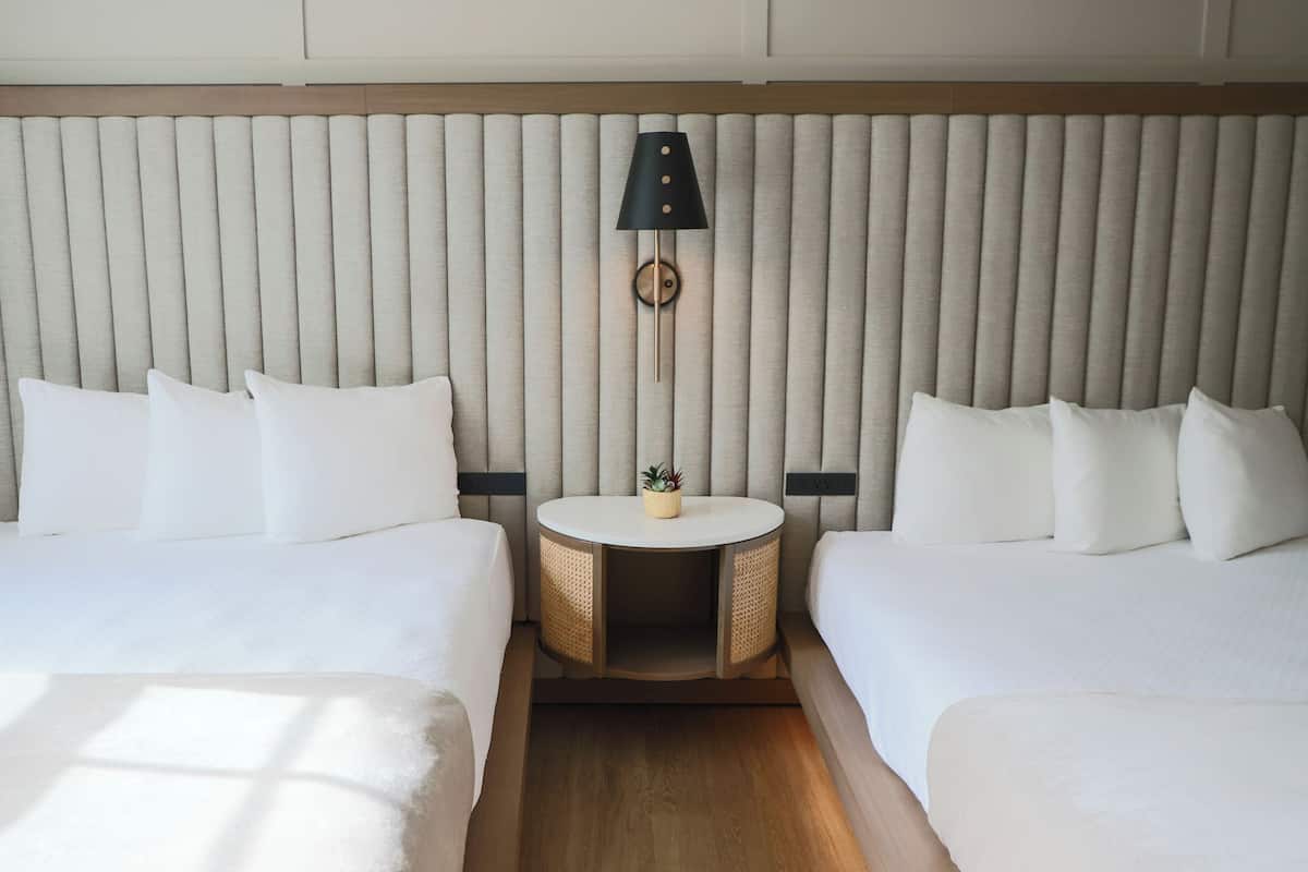 Quarto de hotel com duas camas, uma mesa redonda e uma luminária fixada na parede.
