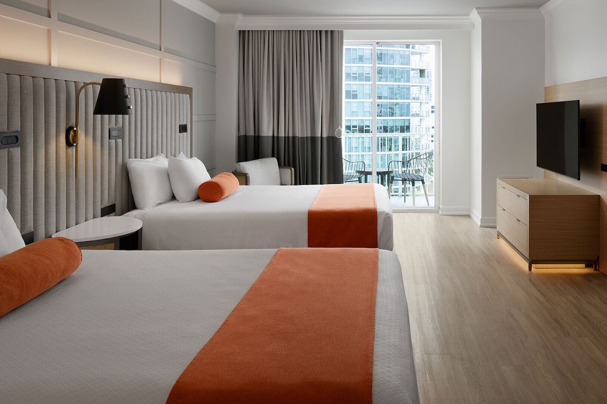 Quarto de hotel com duas camas, televisão, secretária e varanda com vista para a cidade.