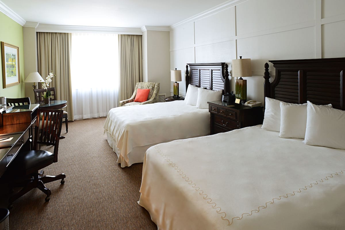 Quarto de hotel com duas camas, uma escrivaninha, uma cadeira, uma luminária e uma janela com cortinas.