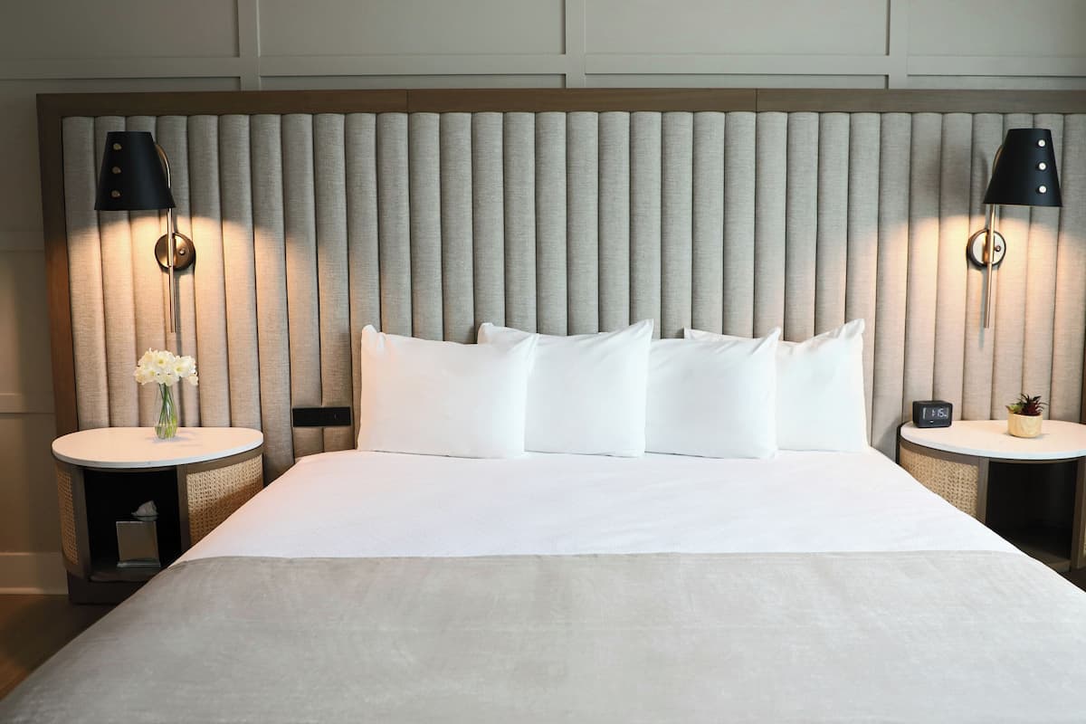 Quarto de hotel com uma cama grande, duas mesas de cabeceira, um relógio fixado na parede e um vaso com flores em uma das mesas.