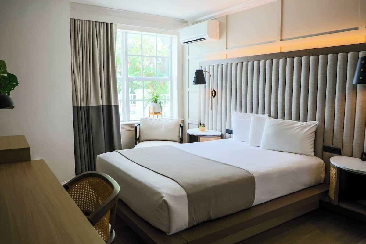 Quarto de hotel com uma cama grande, uma escrivaninha, uma cadeira, uma mesinha com uma planta e uma janela com cortinas.