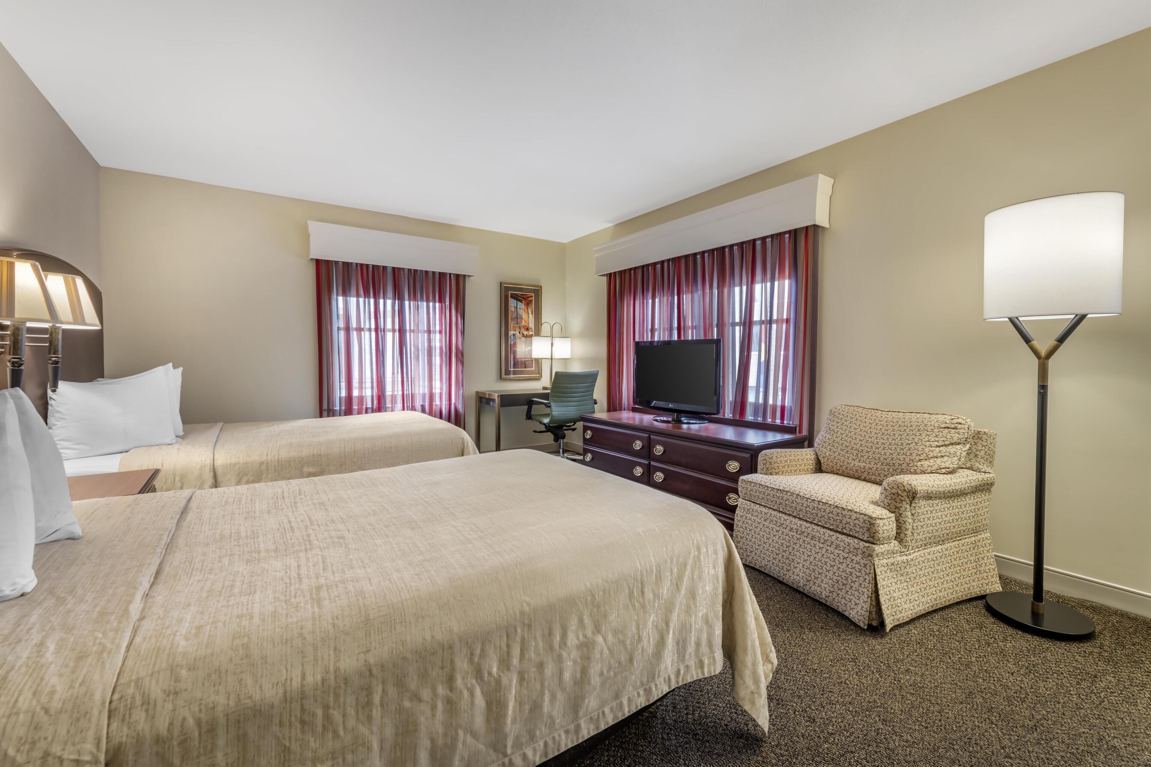 Quarto de hotel com duas camas, uma escrivaninha, televisão, uma cadeira e abajur.