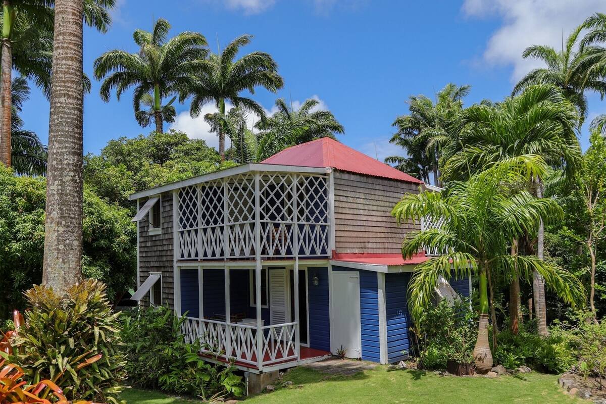 Uma casa de dois andares com telhado vermelho, rodeada por vegetação exuberante e palmeiras.