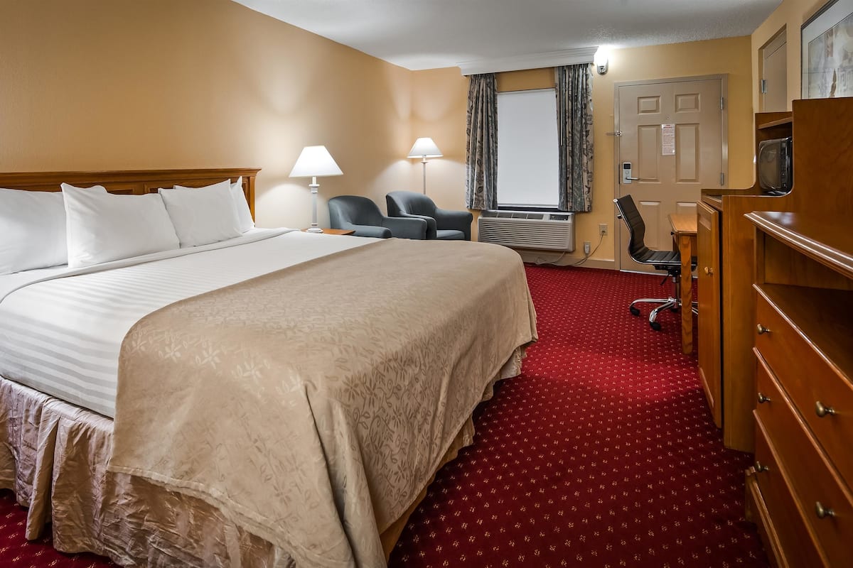 Quarto de hotel com uma cama grande, duas luminárias de cabeceira, uma escrivaninha com luminária, uma cadeira, um guarda-roupa e um tapete vermelho.