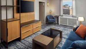 Quarto de hotel moderno com televisão de tela plana na parede, móvel de entretenimento de madeira, sofá azul com almofada laranja, mesa de centro e vista para a cidade pela janela.
