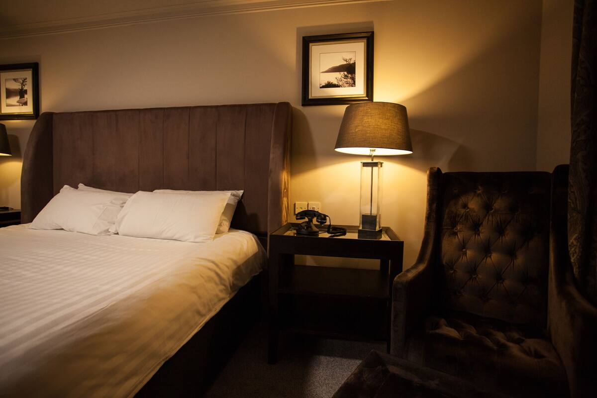 Quarto de hotel com uma cama grande, uma mesa de cabeceira com abajur, uma cadeira e quadros nas paredes.