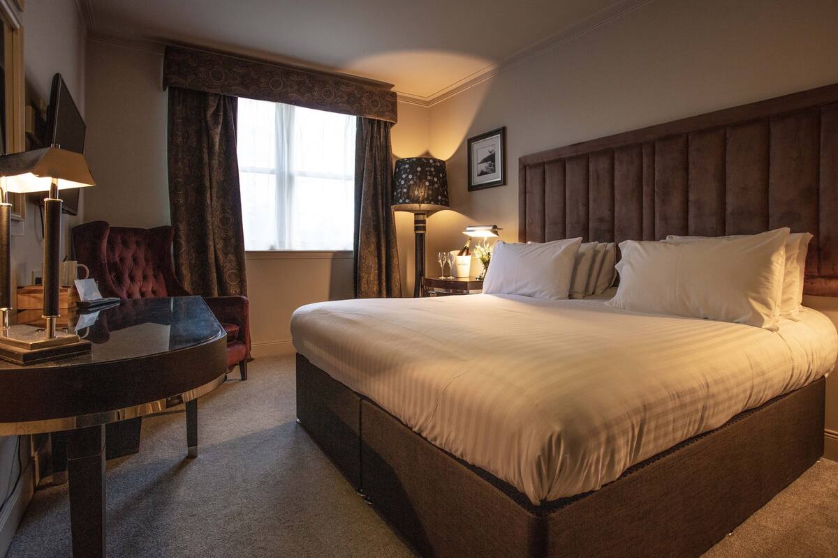 Quarto de hotel com uma cama grande, uma escrivaninha iluminada por uma luminária, uma cadeira e uma janela com cortinas.