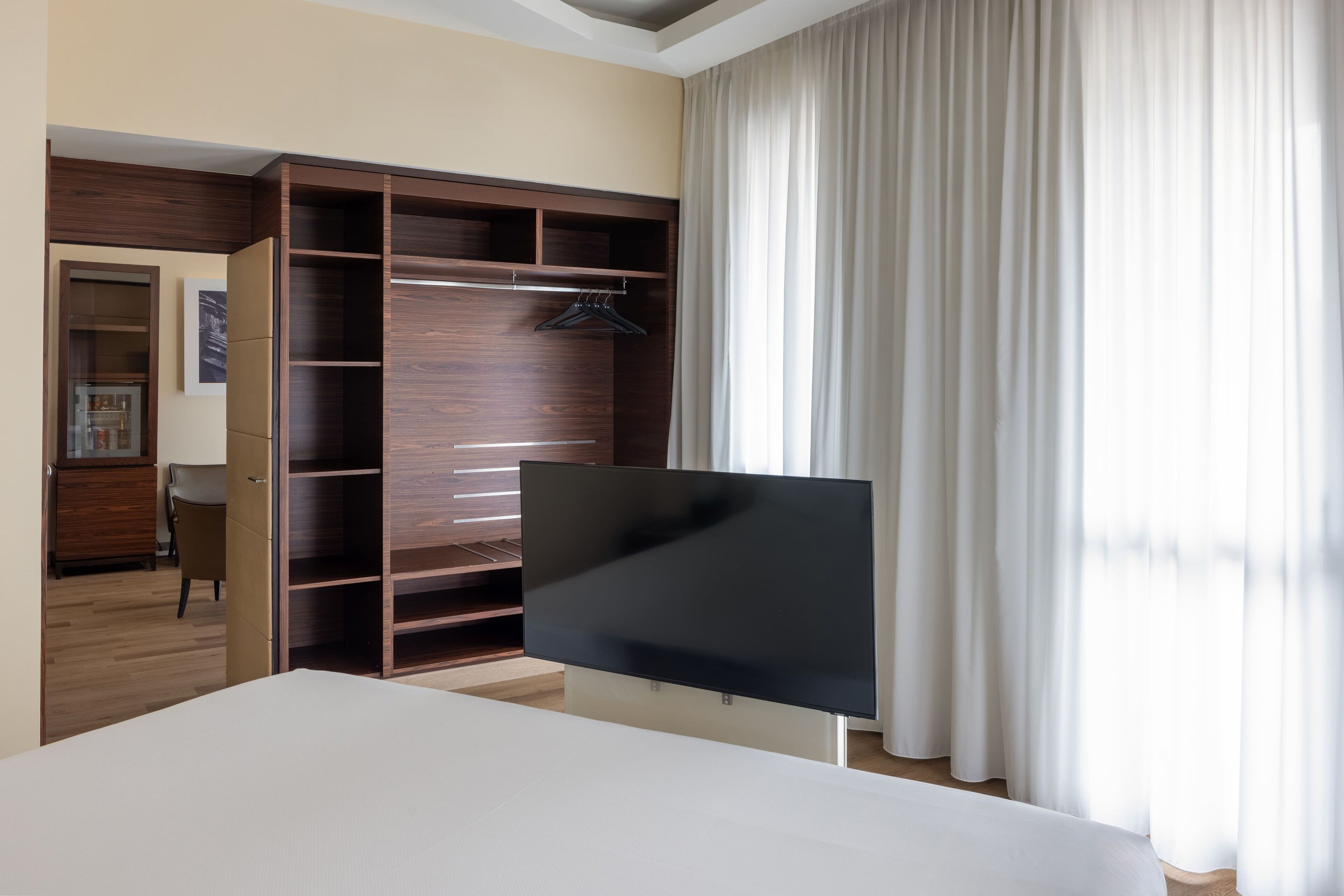Quarto de hotel com cama grande, guarda-roupa com vários compartimentos, televisão sobre suporte e uma janela ampla com cortinas.