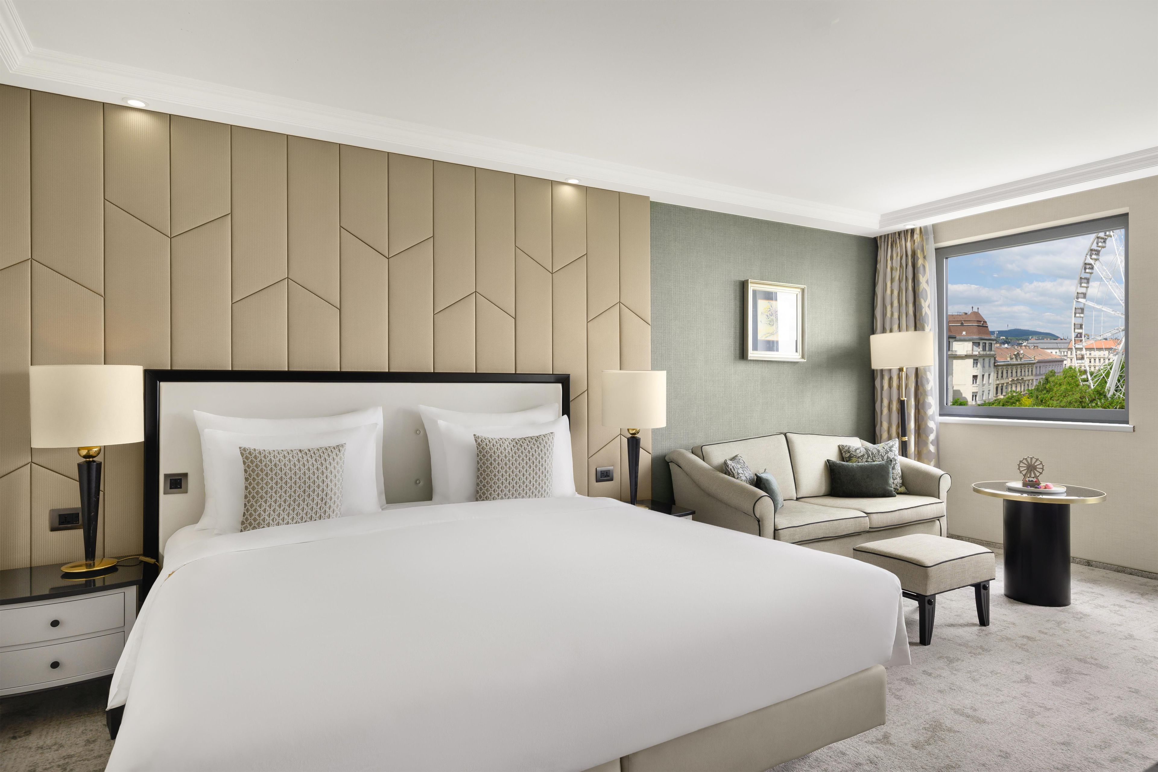 Quarto de hotel com uma cama grande, duas mesas de cabeceira, um sofá, uma mesinha e uma janela vista para uma roda gigante.