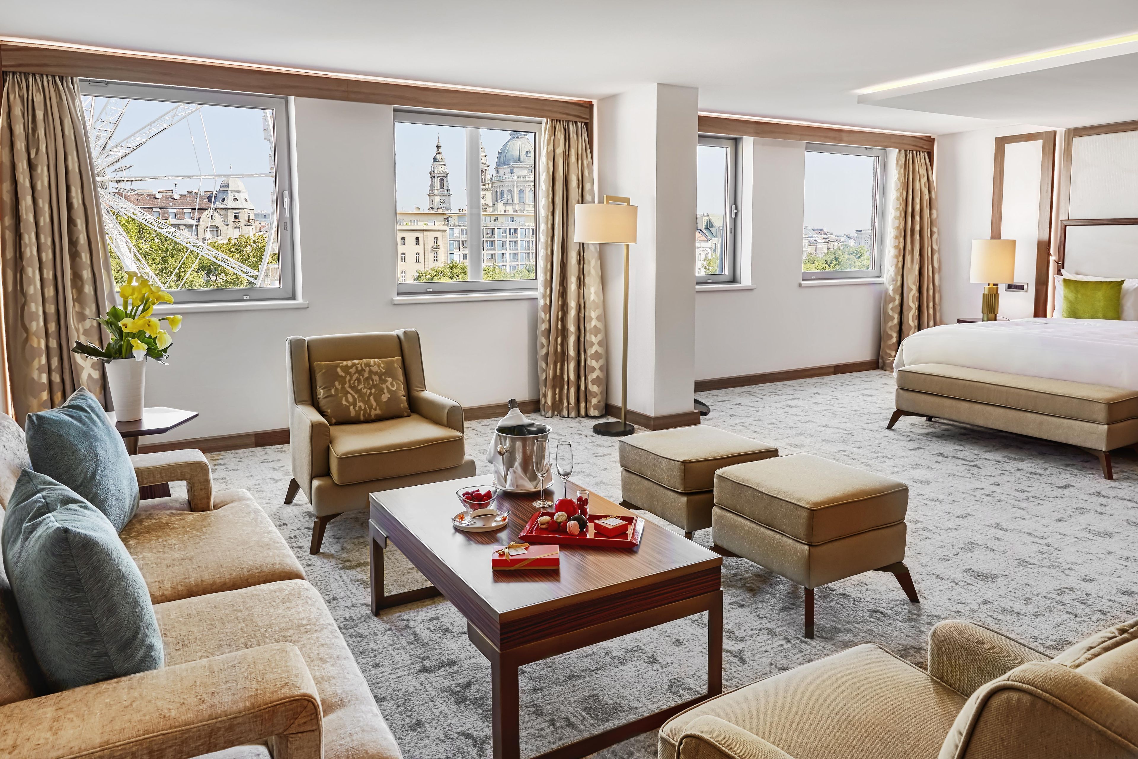Quarto de hotel com uma cama grande, um sofá, uma mesa de centro e vista para a paisagem urbana pelas janelas.