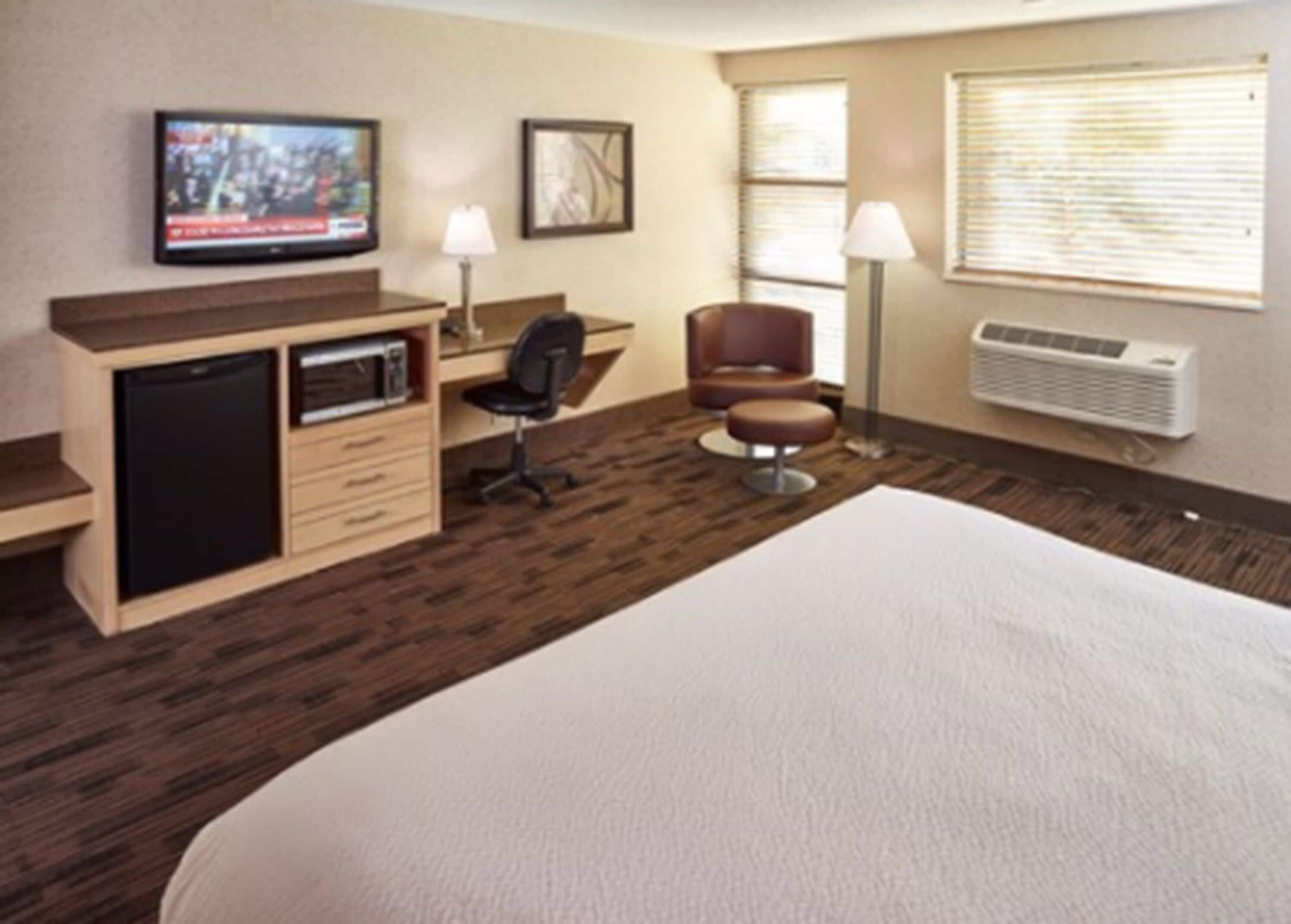 Quarto de hotel com uma cama, uma escrivaninha com cadeira, uma televisão, duas luminárias e um aparelho de ar condicionado.
