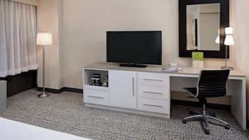 Quarto de hotel com televisão de tela plana, escrivaninha com cadeira, luminária e um espelho grande.